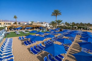 Um resort à beira-mar com fileiras de espreguiçadeiras e guarda-sóis, área de piscina e palmeiras.