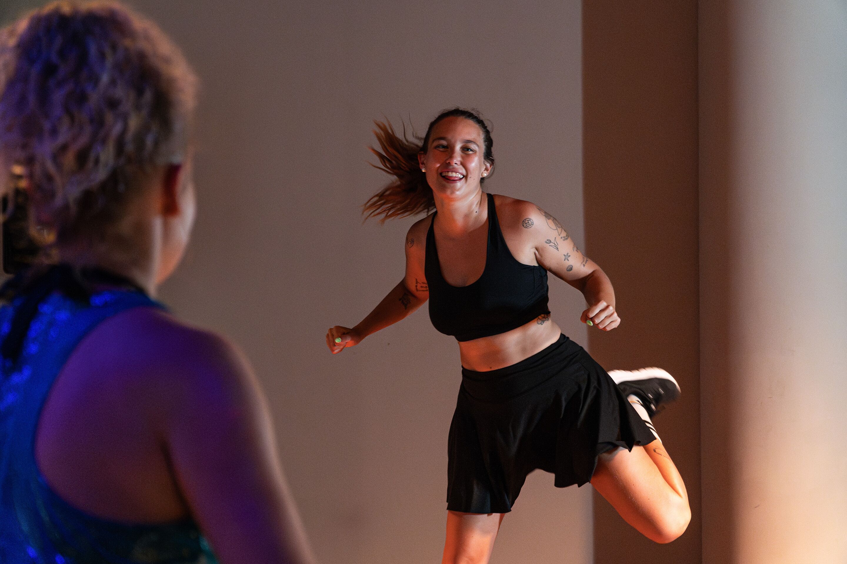 Uma pessoa vestida com roupa de ginástica preta e com tatuagens visíveis, saltando em um ambiente interno.