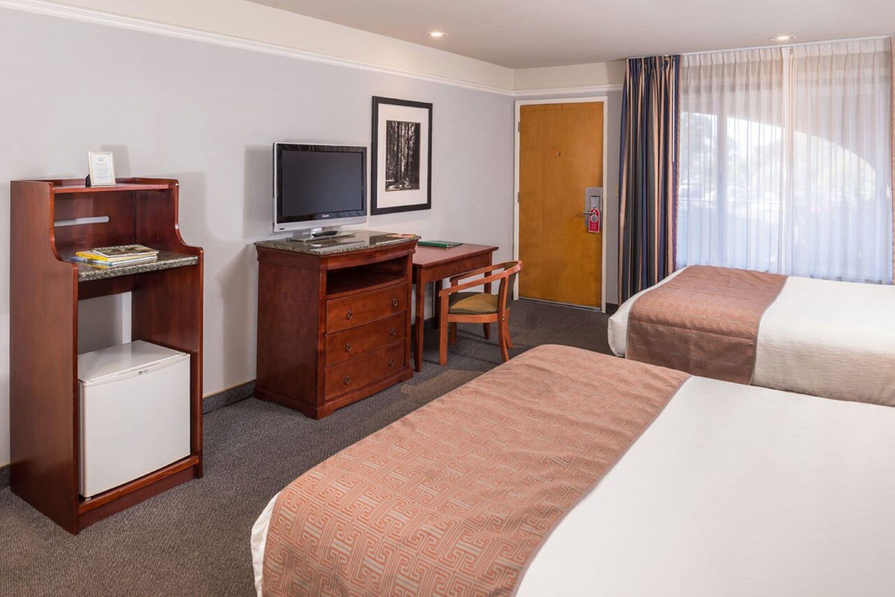 Quarto de hotel com duas camas, uma escrivaninha com computador, um pequeno frigorífico, uma estante e uma porta com cortina.