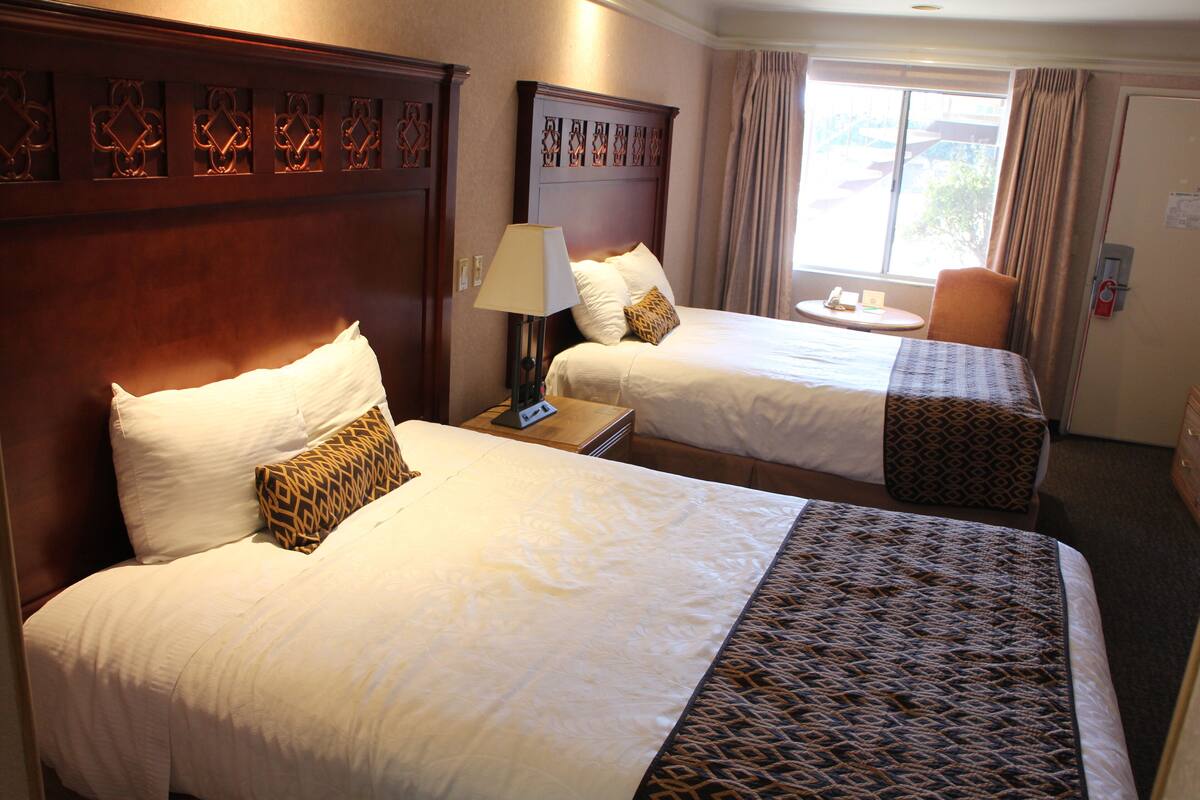 Quarto de hotel com duas camas, mesa de cabeceira, luminária, cadeira e janela com cortinas.