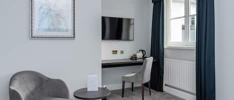 Quarto de hotel moderno com um sofá cinzento, uma mesa redonda, uma escrivaninha com uma TV de tela plana, uma cadeira e uma janela com cortinas escuras.