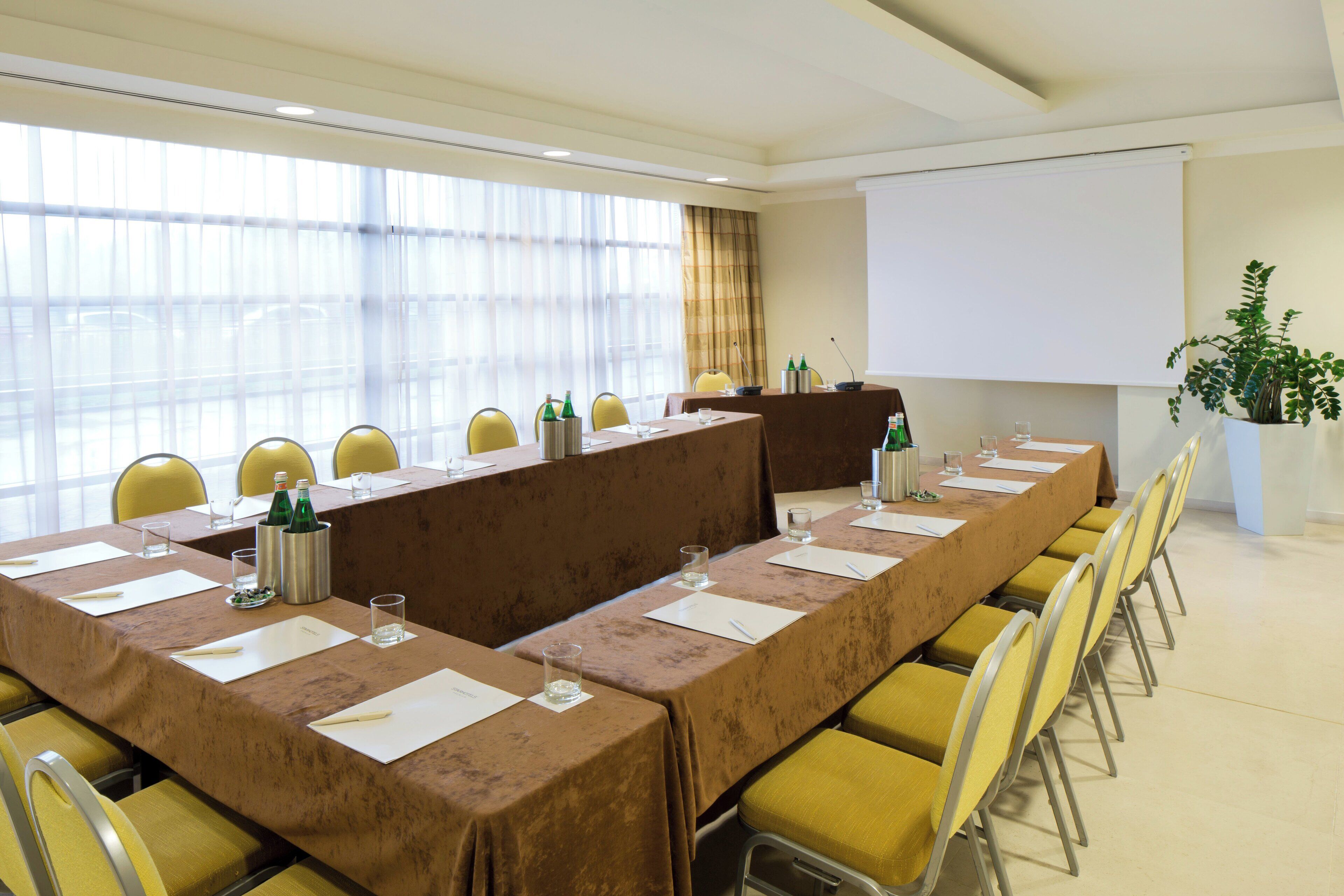 Uma sala de conferências com uma mesa comprida, cadeiras amarelas e uma tela de projetor.