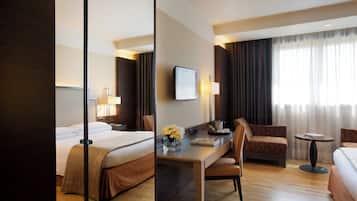 Quarto de hotel com cama, escrivaninha, cadeira, mesa pequena e televisão.