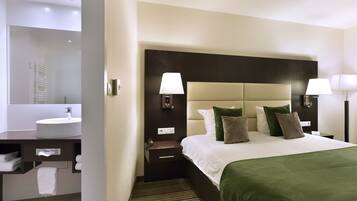 Quarto de hotel com uma cama grande, duas mesas de cabeceira com luminárias, banheiro com pia e espelho, e um porta-toalhas.