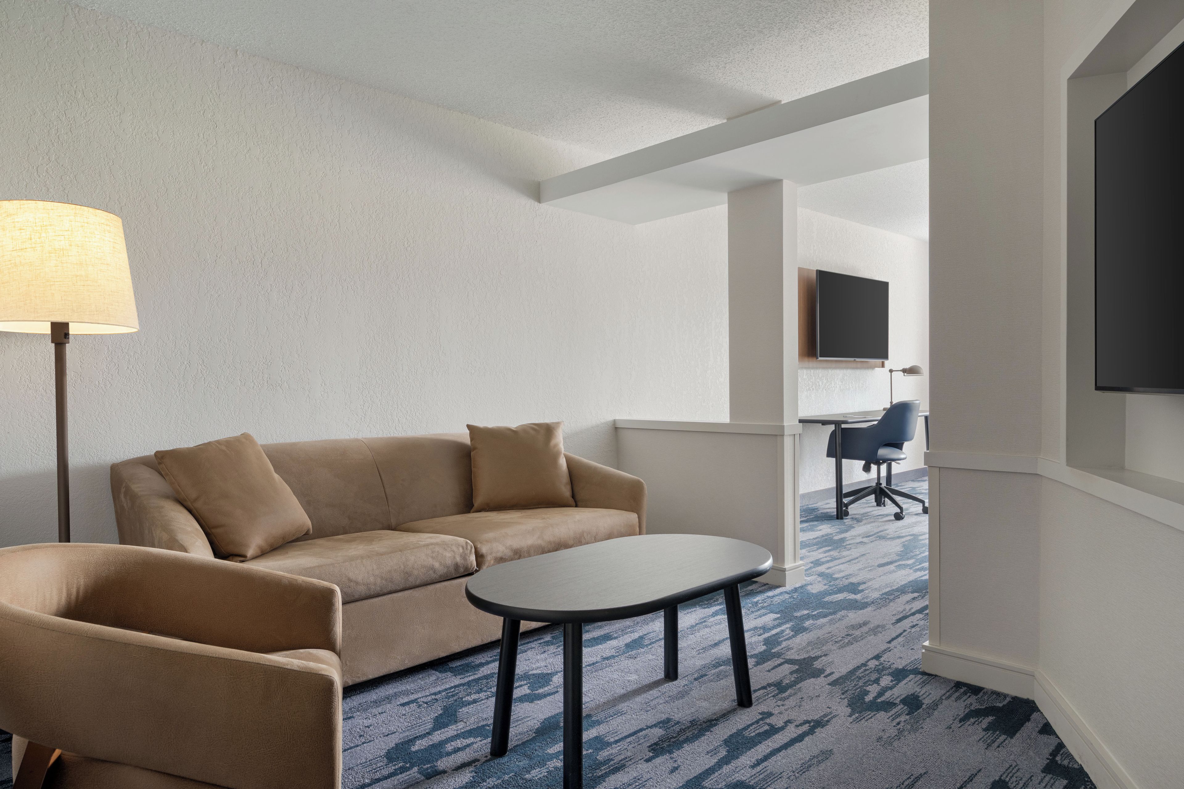 Quarto de hotel com sofá, mesa pequena, luminária, televisão e escrivaninha com cadeira.