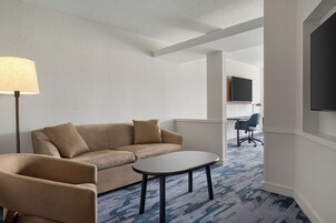 Quarto de hotel com sofá, mesa pequena, luminária, televisão e escrivaninha com cadeira.