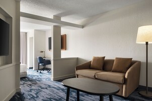 Quarto de hotel com sofá, mesa pequena, luminária, televisão e escrivaninha com cadeira.