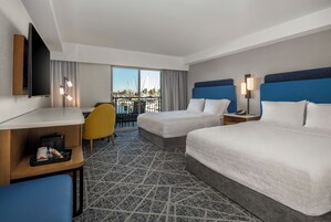 Quarto de hotel com duas camas, uma escrivaninha, uma cadeira, televisão e varanda com vista para barcos.