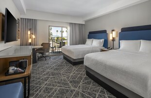 Quarto de hotel com duas camas, uma escrivaninha, uma cadeira, uma televisão e uma varanda com vista.