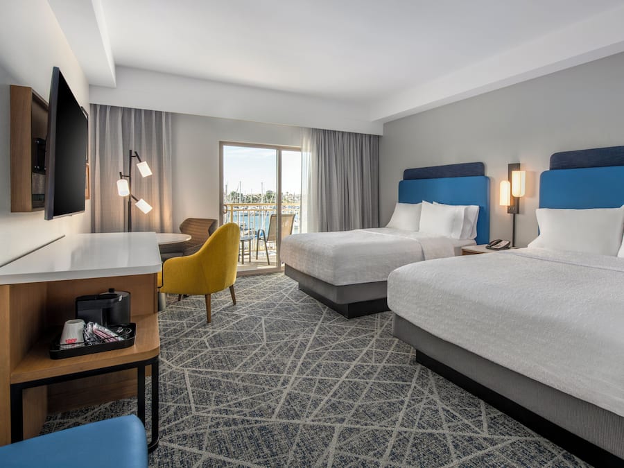 Quarto de hotel com duas camas, uma escrivaninha, uma cadeira, televisão e vista para o porto de iates.