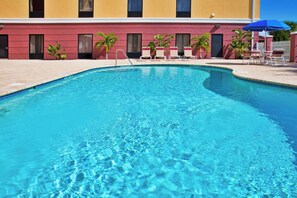 Piscina azul cristalina com escada, rodeada por espreguiçadeiras e guarda-sóis, e um edifício ao fundo.