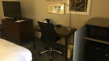 Quarto de hotel com cama, escrivaninha com computador, cadeira, luminária, televisão e minibar.