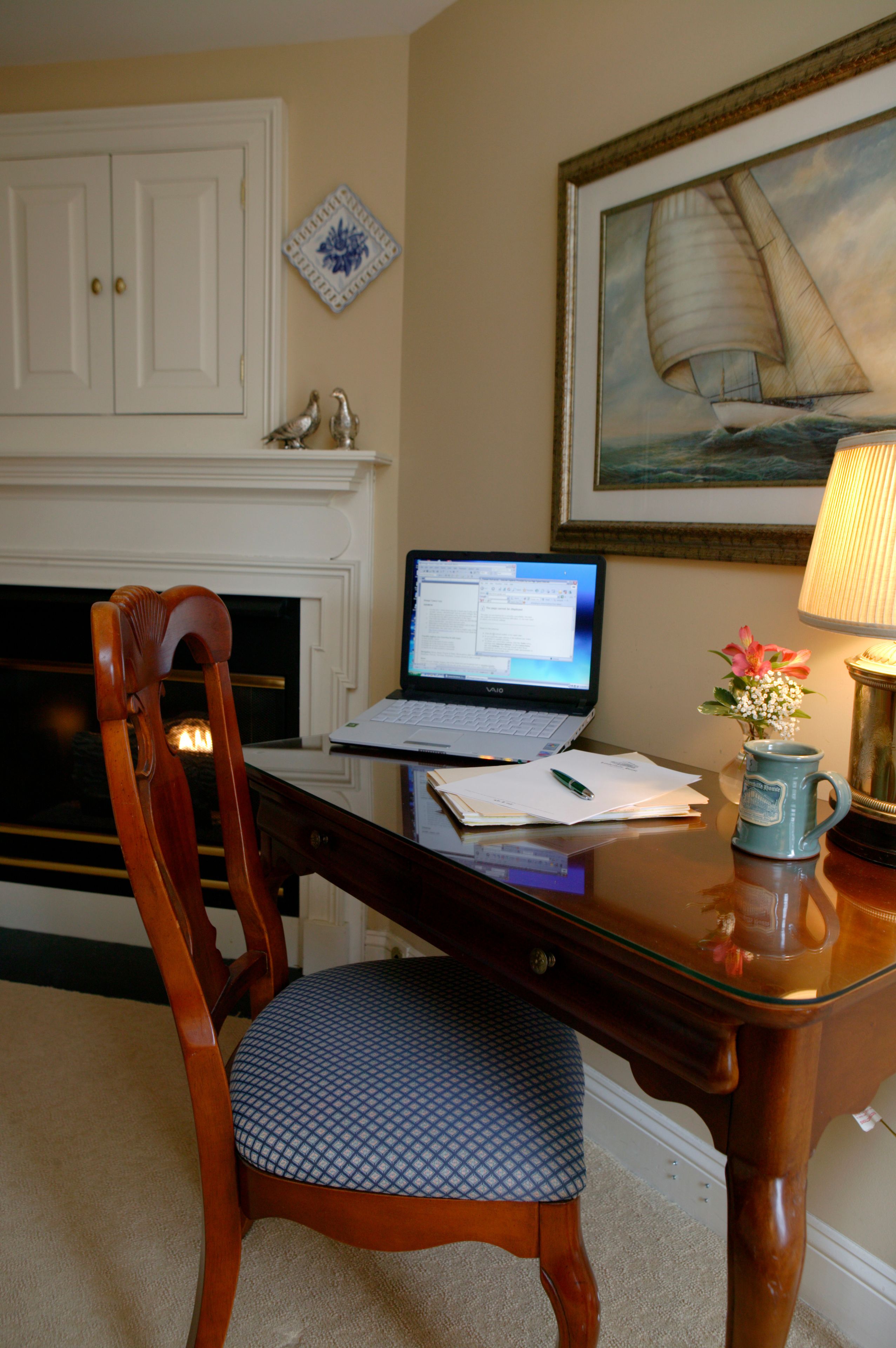 Uma mesa de madeira com um laptop, uma cadeira, um vaso com flores e um quadro emoldurado na parede.