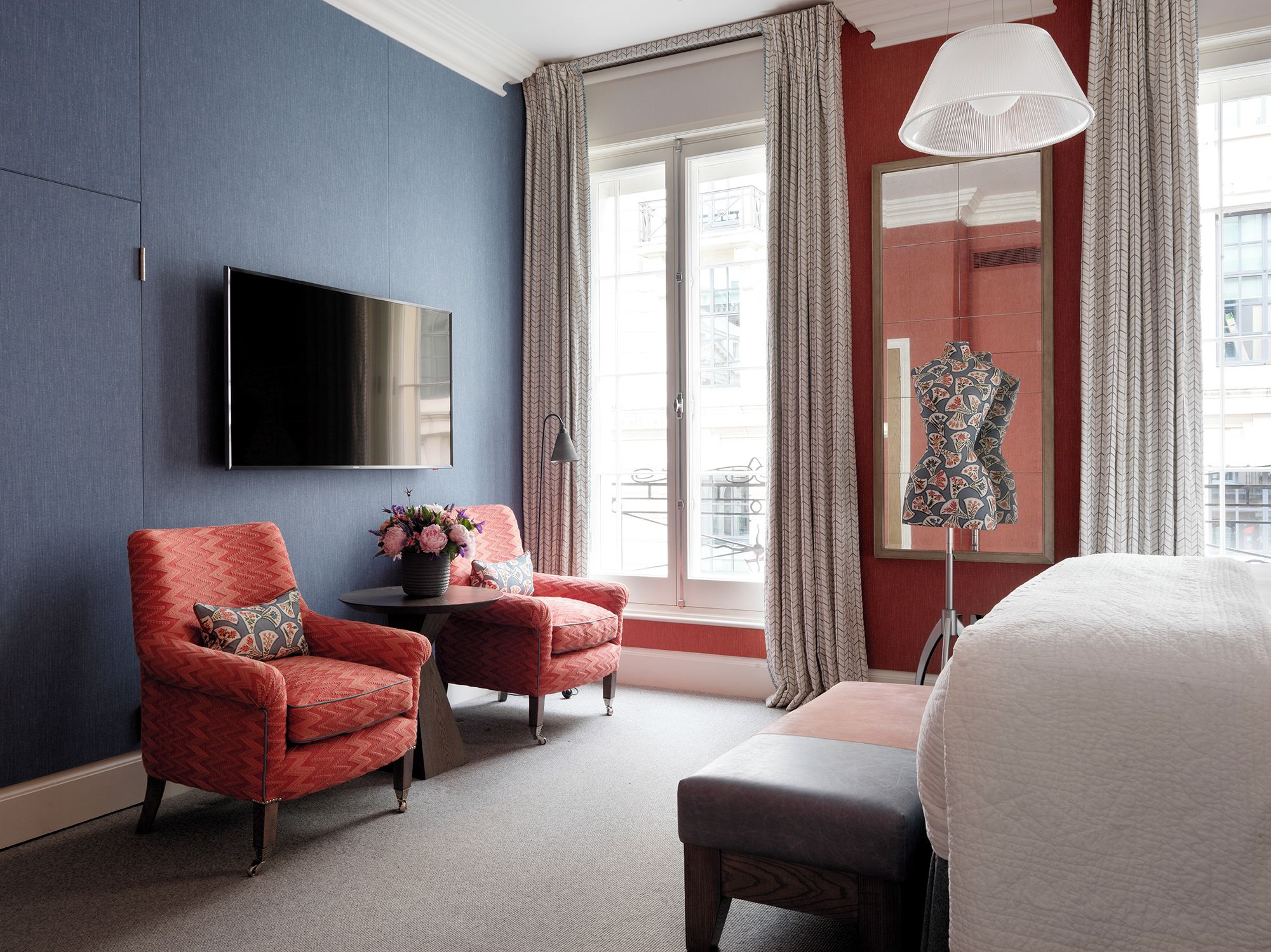 Quarto de hotel com televisão de tela plana, dois poltronas, uma mesinha de centro com um vaso de flores, uma janela grande com cortinas e uma cama com edredom branco.