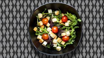 Uma salada em uma tigela com croutons e tomates cereja sobre uma toalha de mesa estampada.