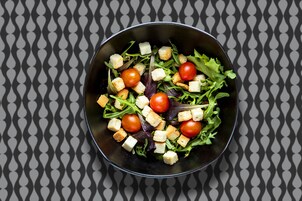 Uma salada em uma tigela com croutons e tomates cereja sobre uma toalha de mesa estampada.