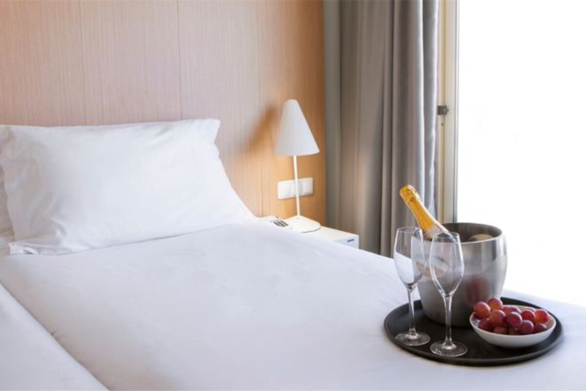 Quarto de hotel com cama, mesa de cabeceira, garrafa de champanhe, copos e uma tigela de uvas.