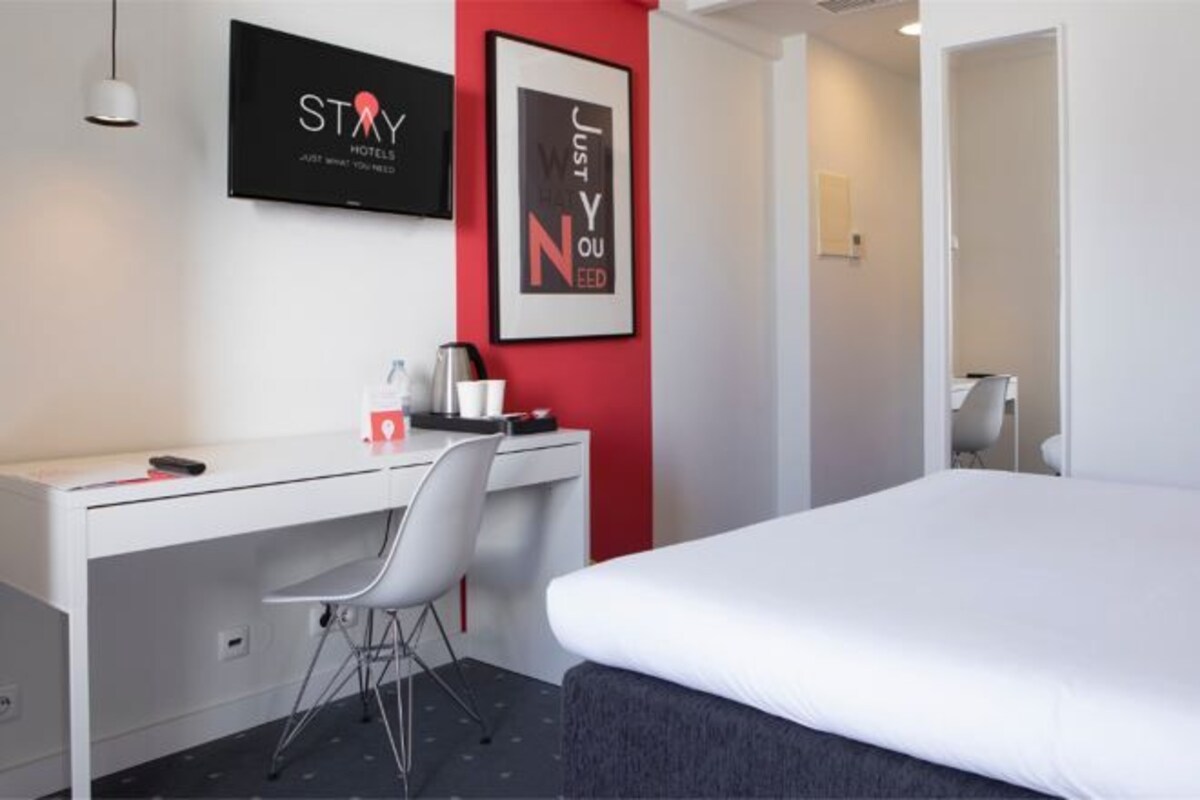 Quarto de hotel com uma cama, uma escrivaninha, uma cadeira, uma cafeteira e um quadro na parede.