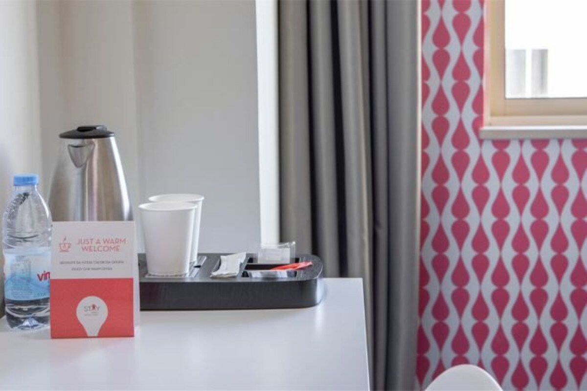 Quarto de hotel com uma mesa branca, um tabuleiro com uma cafeteira, uma garrafa de água, uma chávena e um cartão de boas-vindas.