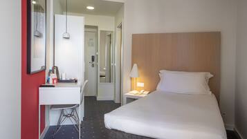 Quarto de hotel com uma cama, uma escrivaninha com cadeira, um abajur e um espelho.