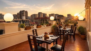Terraço no telhado com mesas preparadas para refeições, plantas em vasos e vista para a cidade ao pôr do sol.