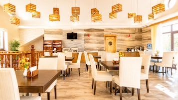 Uma área de refeições de hotel com mesas de madeira, cadeiras brancas e luminárias pendentes de madeira.