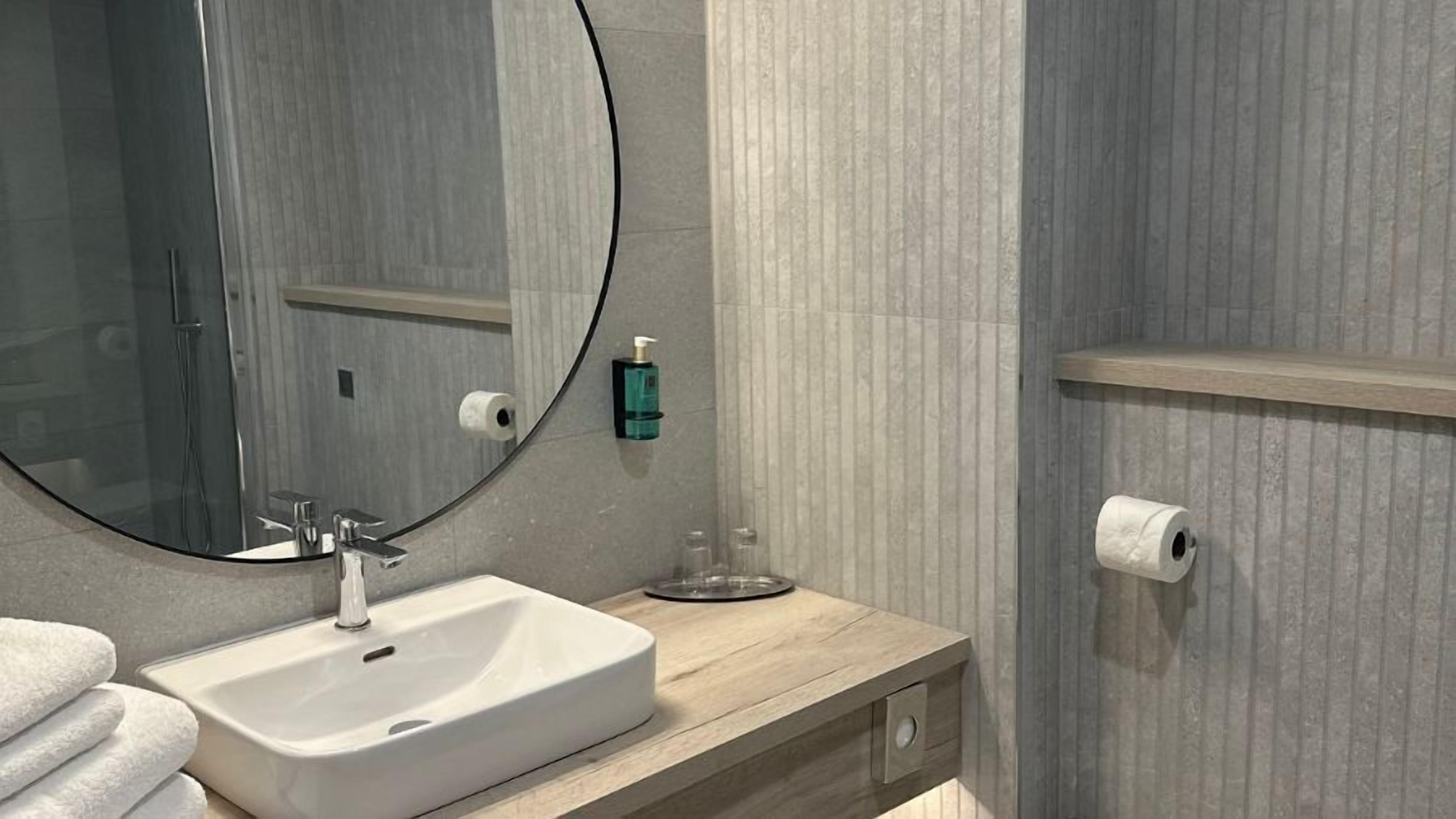 Casa de banho com lavatório branco, espelho redondo e suporte para papel higiénico.