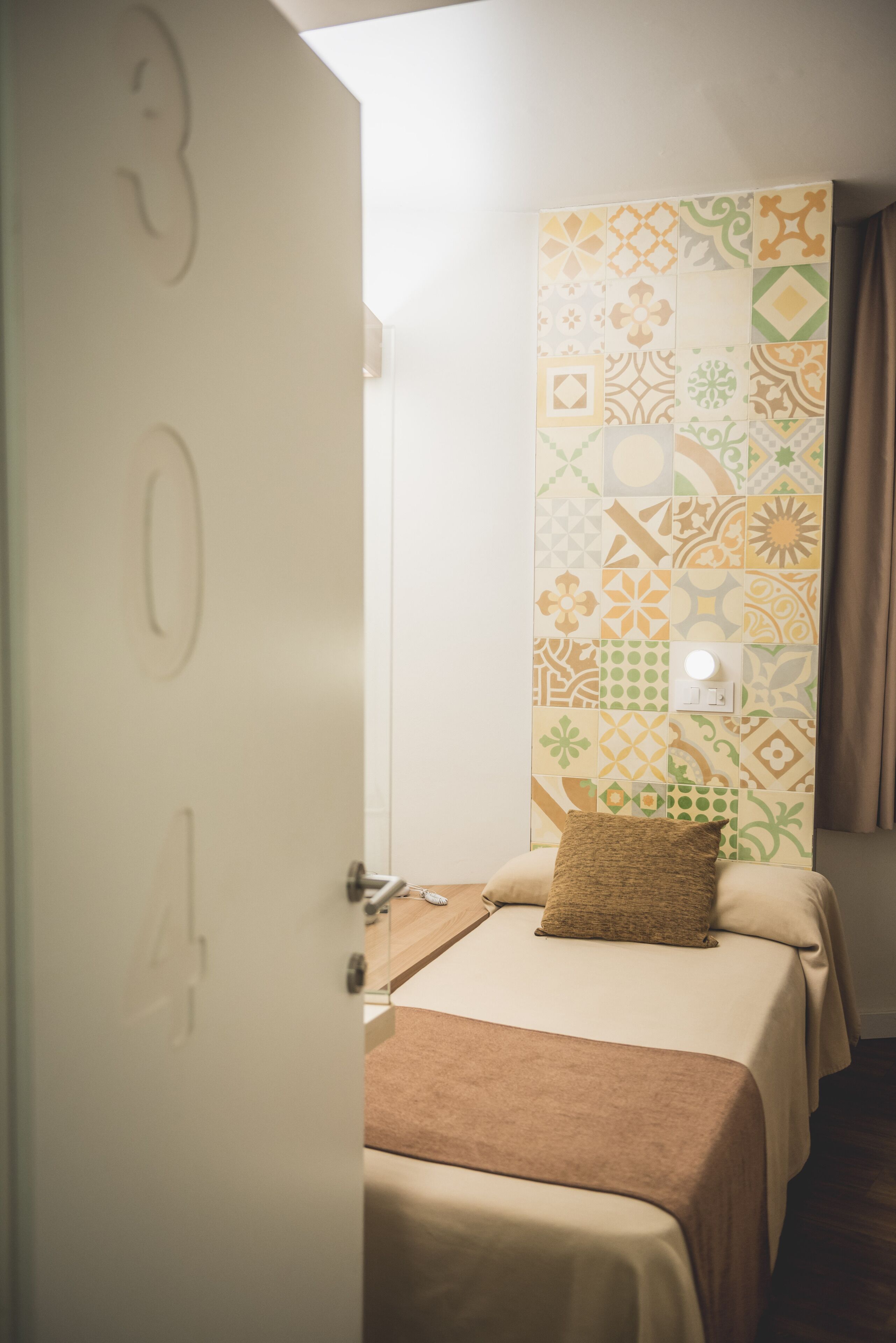 Quarto de hotel com uma cama, mesa de cabeceira, rodapé e uma porta com número.