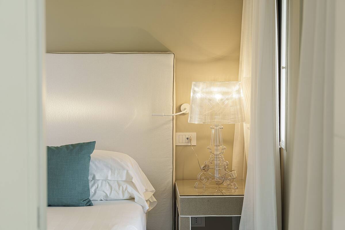 Quarto de hotel com cama, mesa de cabeceira, luminária e janela com cortinas.