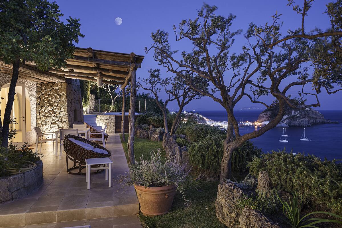 Um pátio com área de estar externa, parede de pedra e vista para o mar à noite.