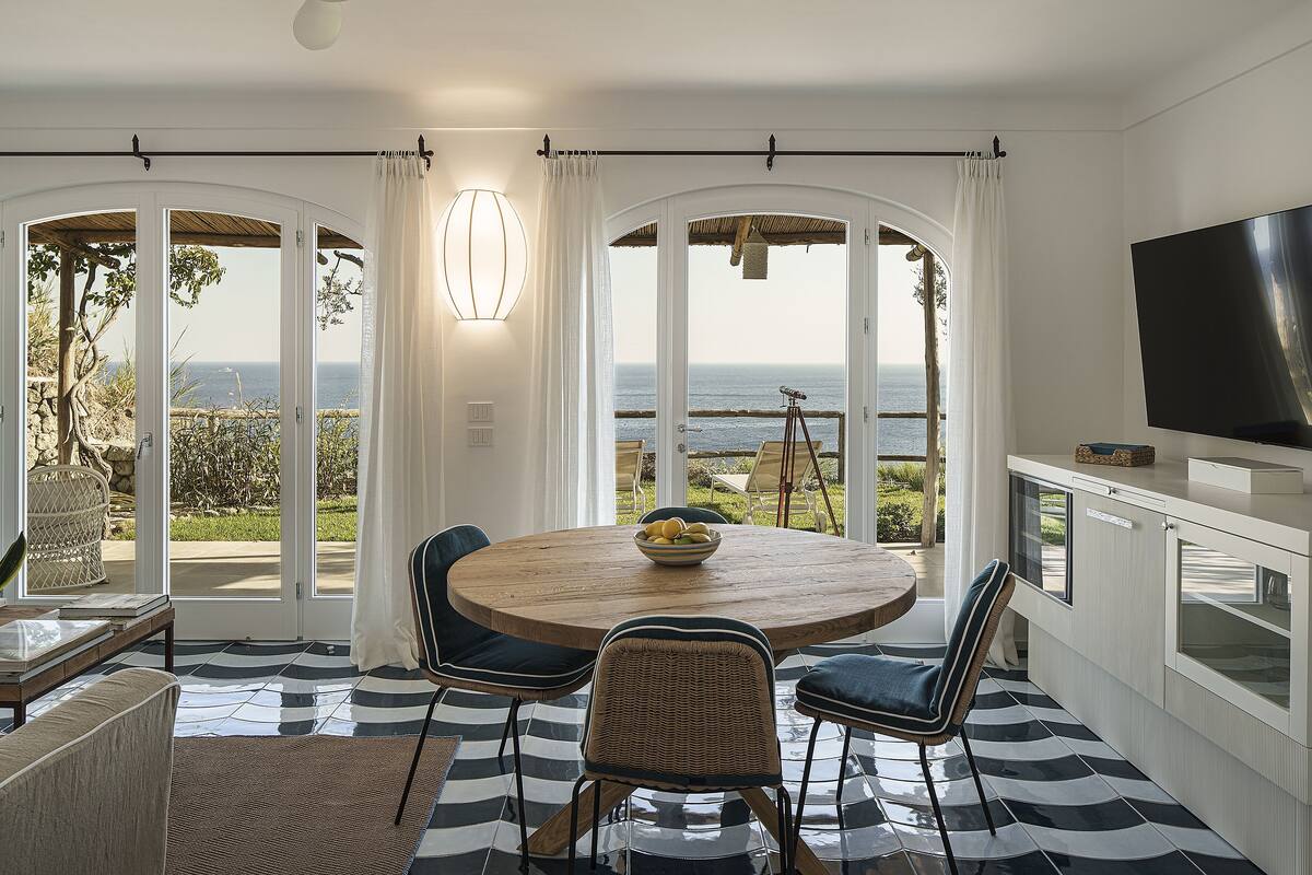 Uma área de refeições com uma mesa redonda, cadeiras e uma tigela de frutas. Há janelas amplas com cortinas, uma TV de tela plana e vista para o mar.