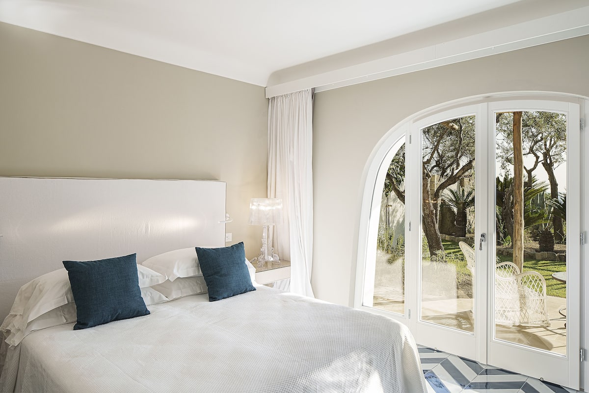 Quarto com cama, mesa de cabeceira, abajur e vista para um pátio externo com cadeiras e uma mesa.
