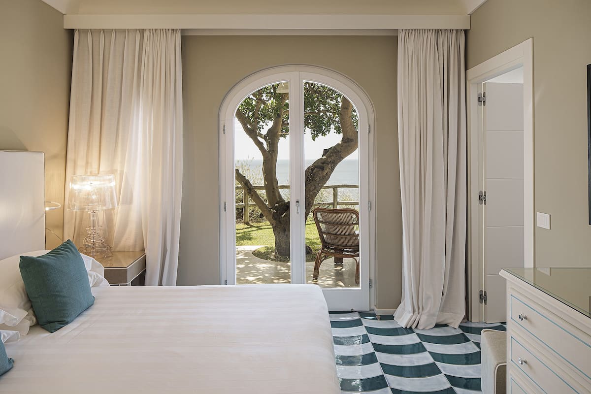 Quarto com uma cama grande, vista do exterior por uma porta de vidro, uma cadeira do lado de fora e uma cômoda com gavetas.