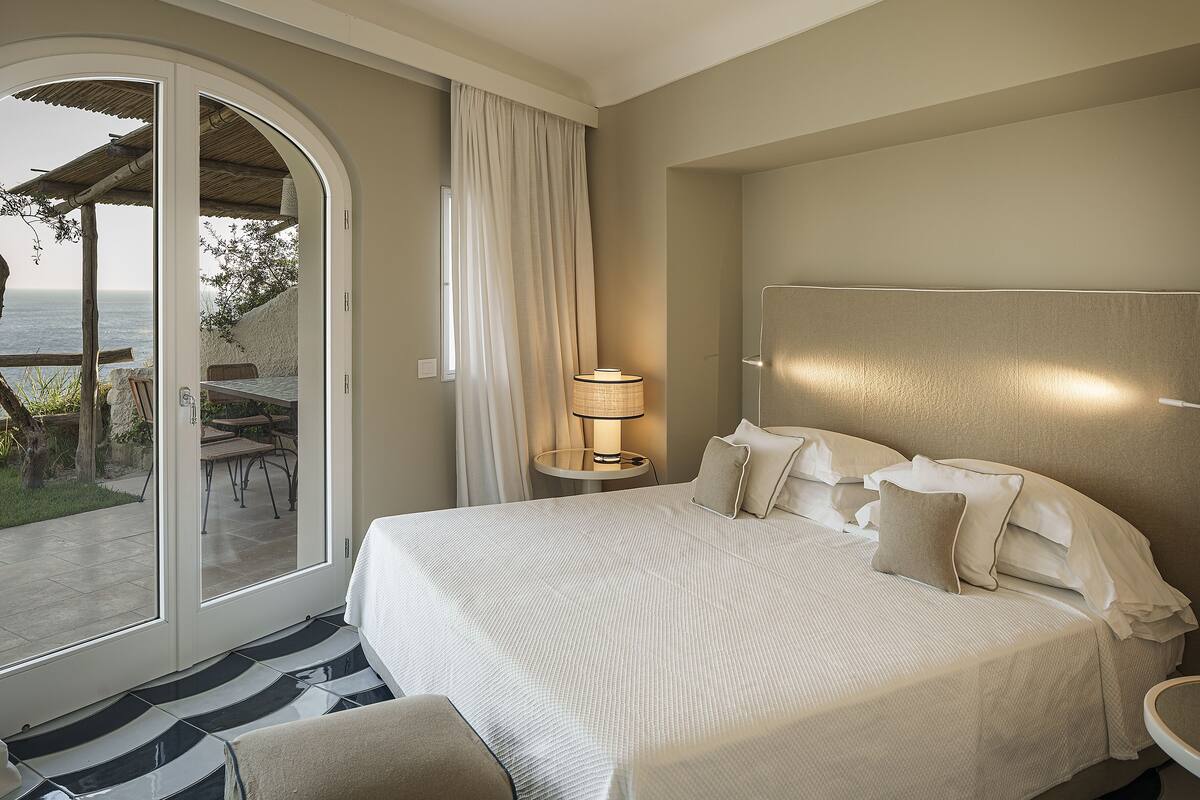 Quarto de hotel com cama grande, mesa de cabeceira com abajur, vista para o mar através de uma porta de vidro e varanda com mobiliário exterior.