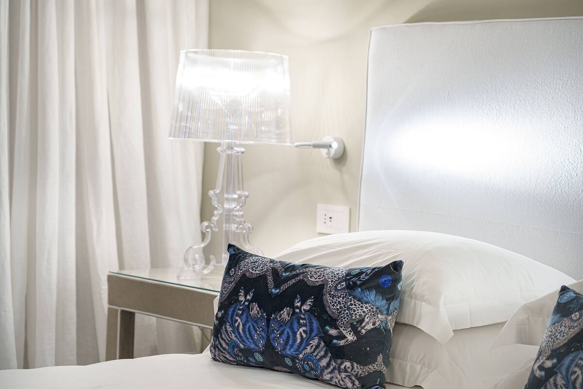 Uma cama bem arrumada com roupa de cama branca, uma luminária de vidro transparente em uma mesa de cabeceira e um travesseiro estampado.