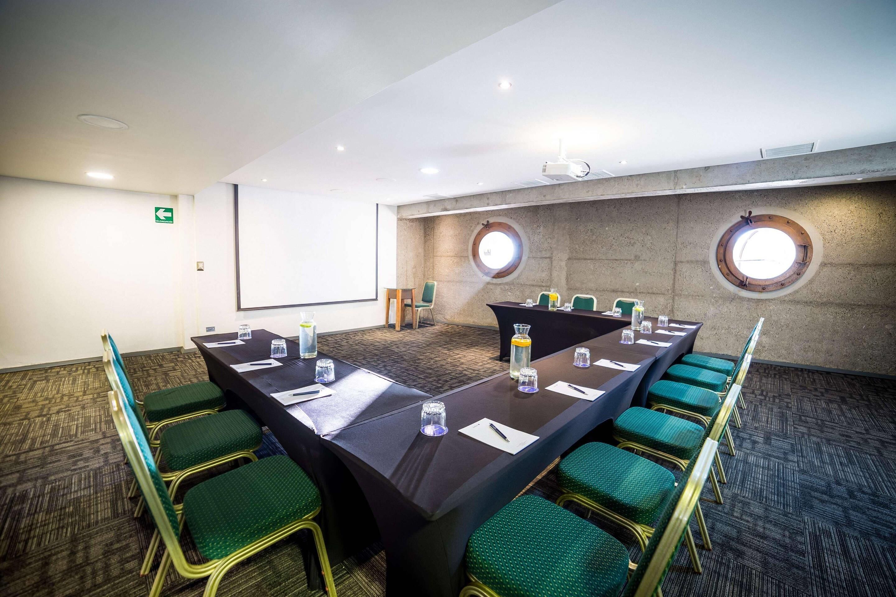 Uma sala de conferências com uma mesa comprida, cadeiras verdes e um projetor.