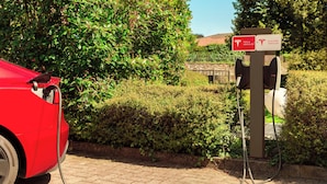 Uma estação de carregamento Tesla vermelha, com placa indicando que é para Teslas e veículos elétricos, rodeada por vegetação e uma área pavimentada.