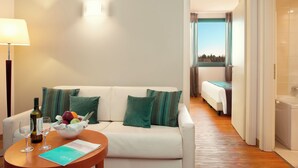 Quarto de hotel com sofá branco, mesa de apoio de madeira com uma garrafa de vinho e copos, uma fruteira, e vista para uma varanda com uma cama.