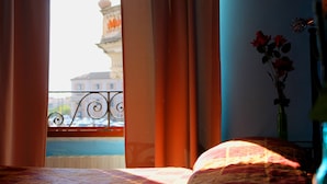 Quarto com cama, janela com cortinas, vaso com flores e vista para edifícios.