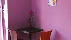 Uma pequena mesa com um vaso de flores e duas cadeiras, uma laranja e outra cinza, em um quarto com paredes roxas.