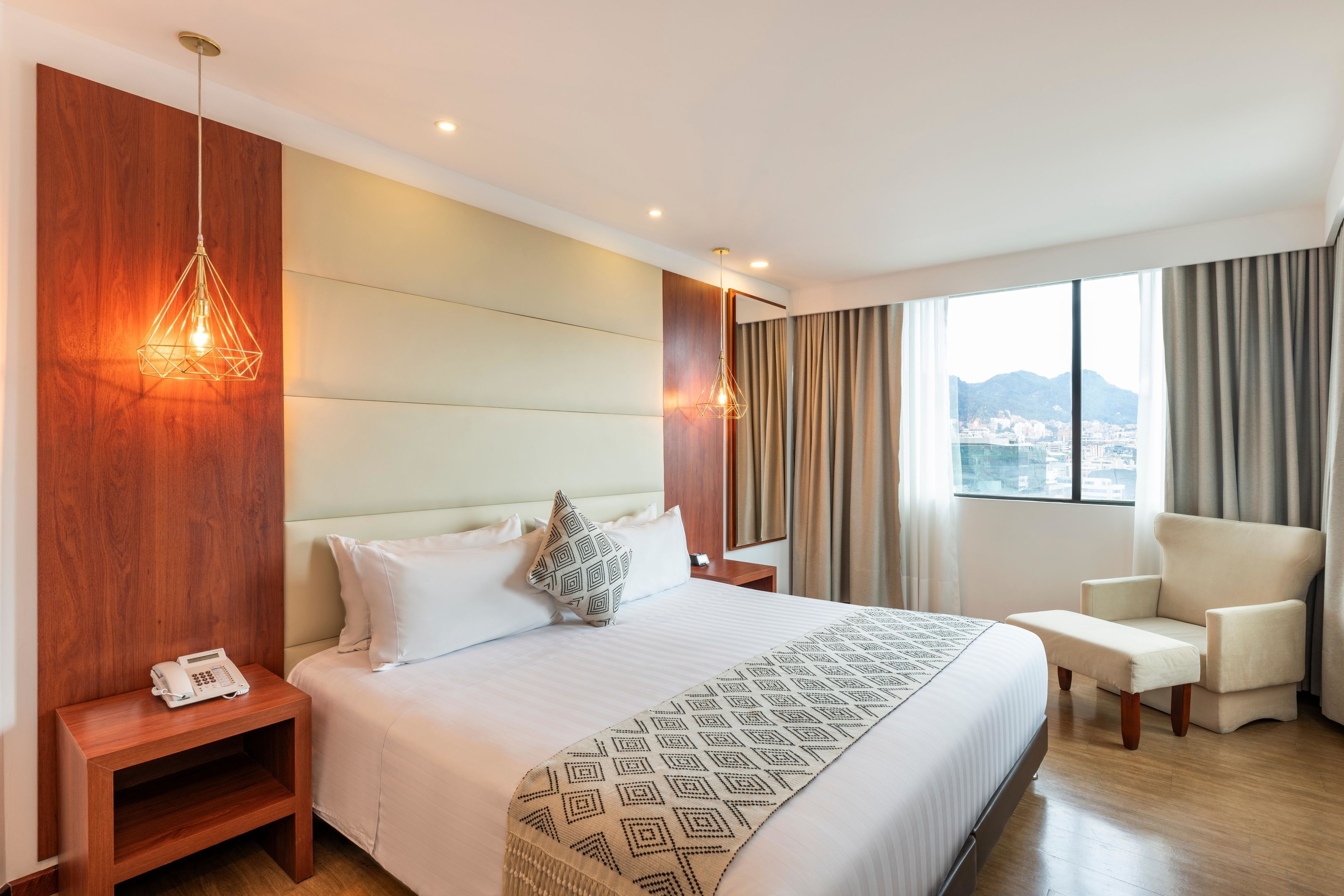 Quarto de hotel com uma cama grande, mesa de cabeceira com telefone, uma cadeira e vista para a cidade através da janela.