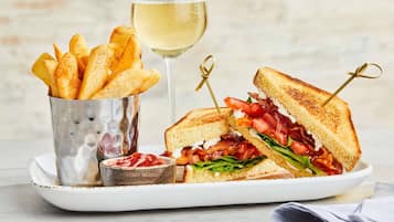 Um prato com um sanduíche grelhado, um copo de vinho branco e um recipiente com batatas fritas.
