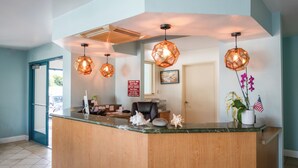 Uma mesa de recepção com balcão de madeira, três luminárias pendentes, um quadro na parede e uma placa indicando o horário de check-in e expediente.