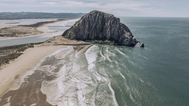 Paisagem costeira com uma formação rochosa proeminente, praia arenosa e mar calmo.