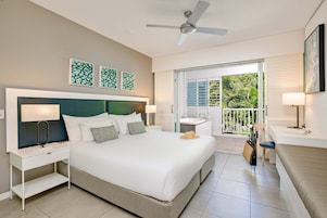 Quarto de hotel moderno com uma cama grande, ventilador de teto, varanda com vista para áreas verdes e uma escrivaninha com cadeira.