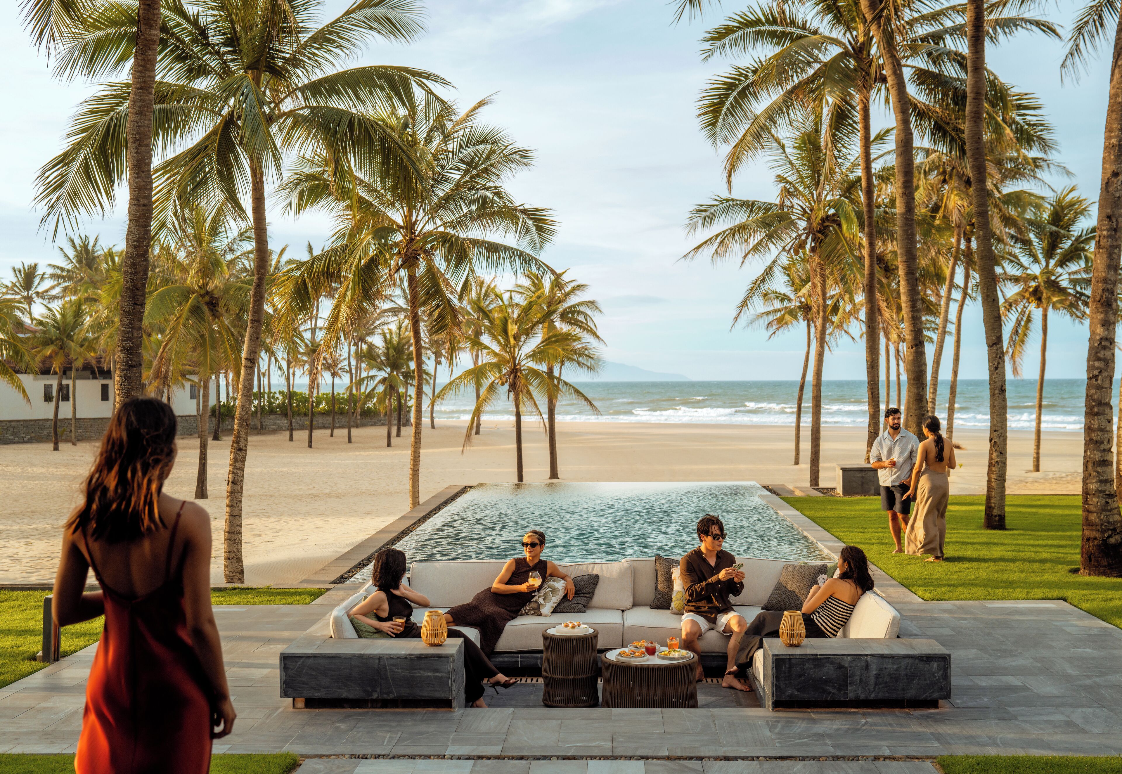 Área à beira da piscina com assentos, palmeiras e vista para a praia.
