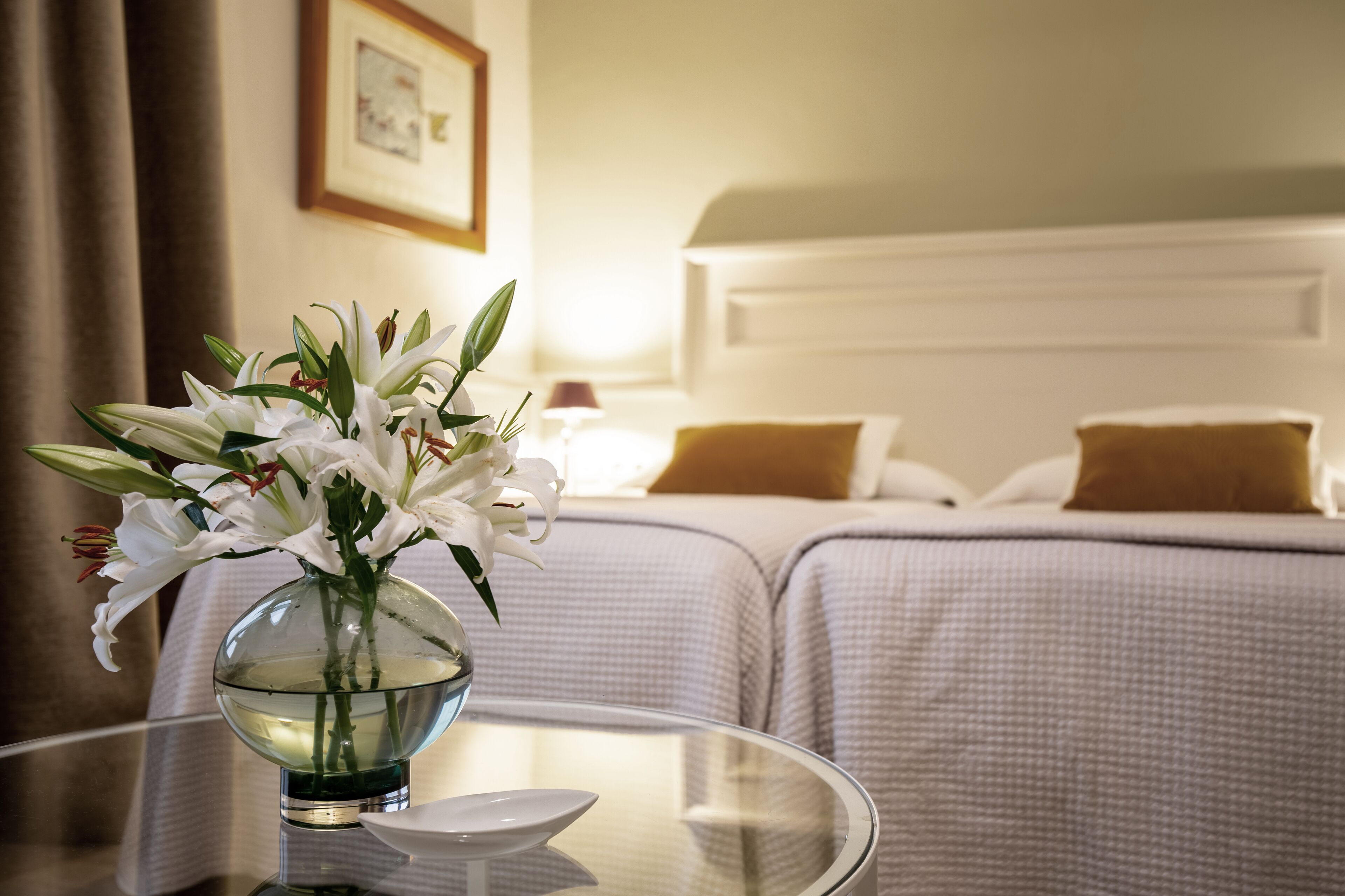 Quarto de hotel com uma cama, uma mesinha de cabeceira com um vaso de flores e um quadro pendurado na parede.