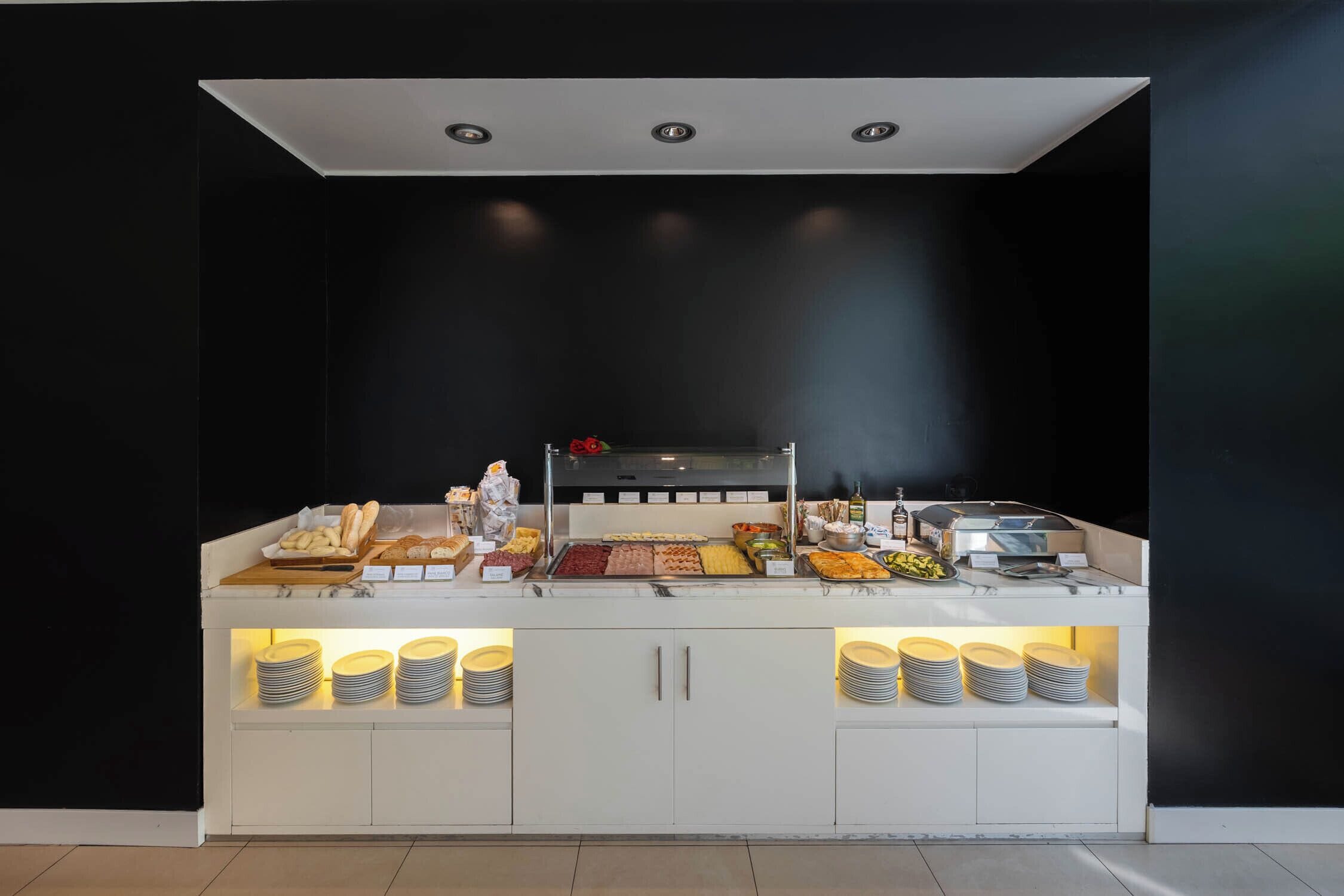 Buffet com diversos alimentos e acompanhamentos, incluindo pão, doces e bebidas, em frente a uma parede escura com balcão branco e pratos iluminados.
