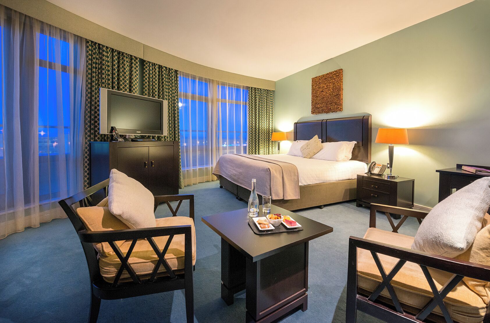Quarto de hotel com uma cama, duas cadeiras, uma escrivaninha com abajur, televisão e minibar.
