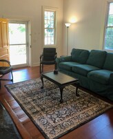 Uma sala de estar com um sofá verde, uma poltrona preta, um tapete estampado e uma mesa.